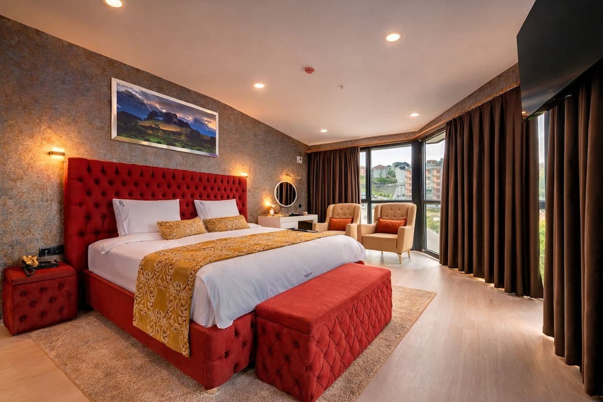 Quarto de hotel moderno com uma cama grande, cabeceira vermelha, vista da cidade e varanda com cortinas.