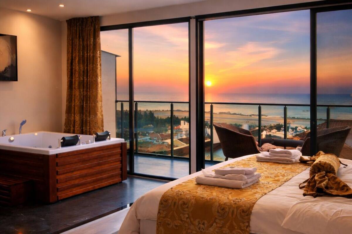 Quarto de hotel moderno com uma cama grande, banheiro com banheira e varanda com vista para o pôr do sol.