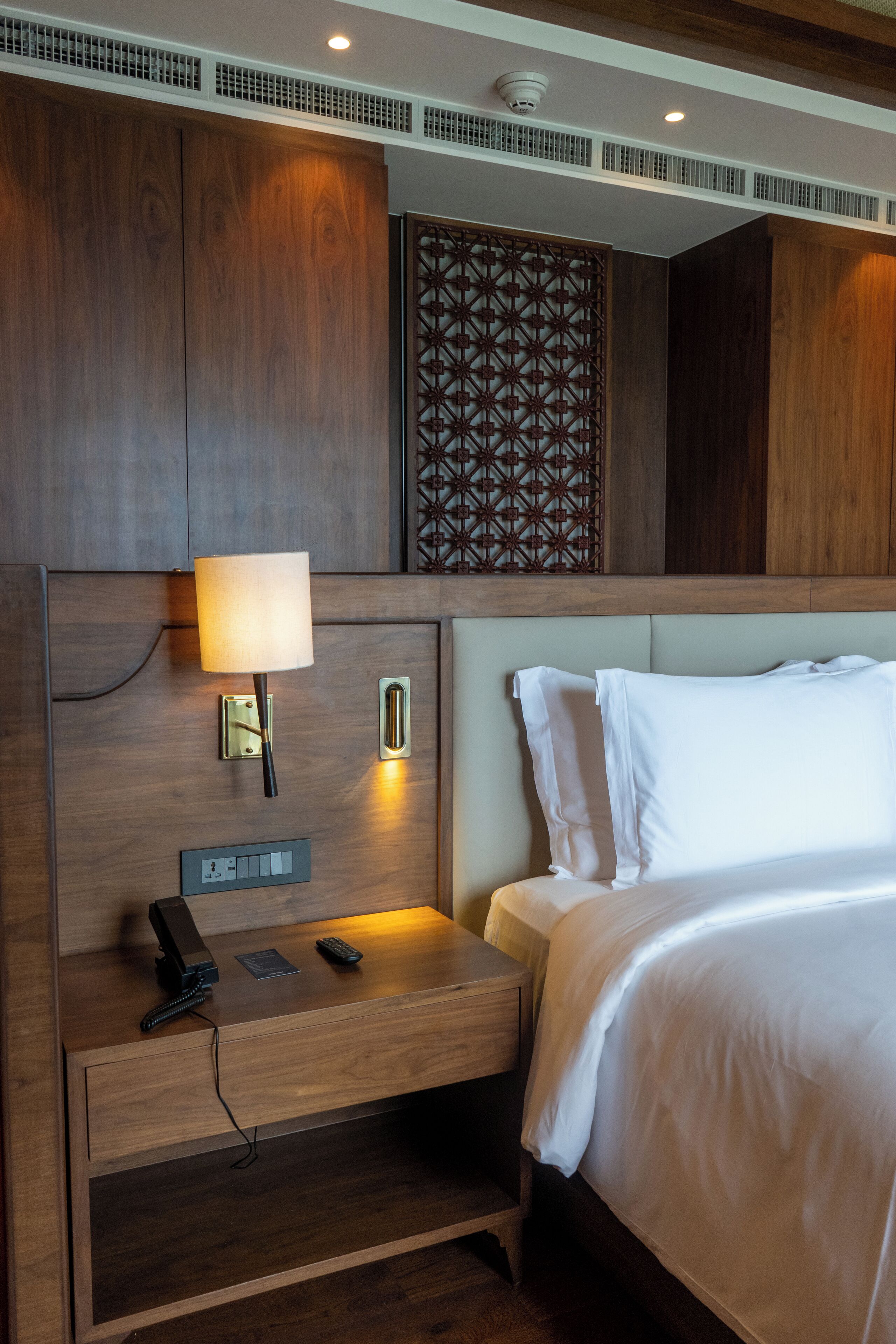 Quarto de hotel com cabeceira de madeira, mesa de cabeceira com luminária, controle remoto e tomada.