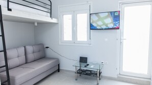 Um quarto com uma cama de solteiro, um sofá, uma mesa de vidro com um controle remoto e uma TV fixada na parede.