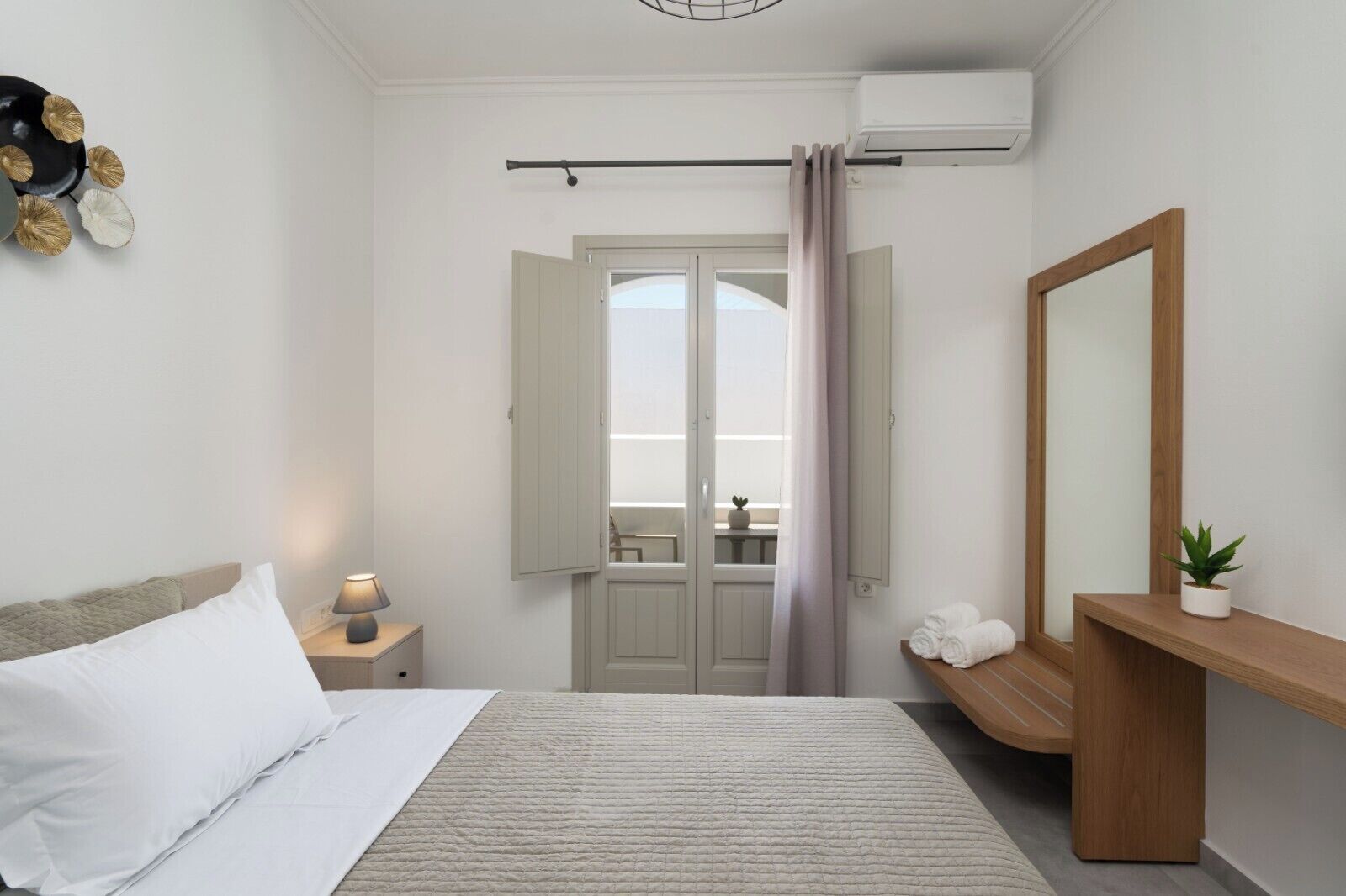 Quarto com cama, luminária de cabeceira, console de madeira com planta, espelho e vista para uma varanda através de uma porta aberta.