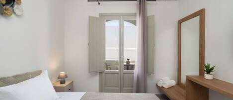 Quarto com cama, luminária de cabeceira, console de madeira com planta, espelho e vista para uma varanda através de uma porta aberta.