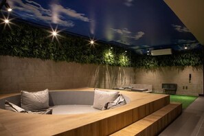 Uma área de relaxamento moderna em ambiente interno, com um banco de madeira curvo, um pequeno espaço de estar e um painel de fundo com plantas artificiais verdes.