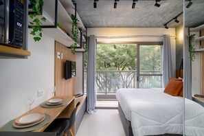 Quarto moderno com cama, escrivaninha com pratos, televisão e varanda com cortinas.