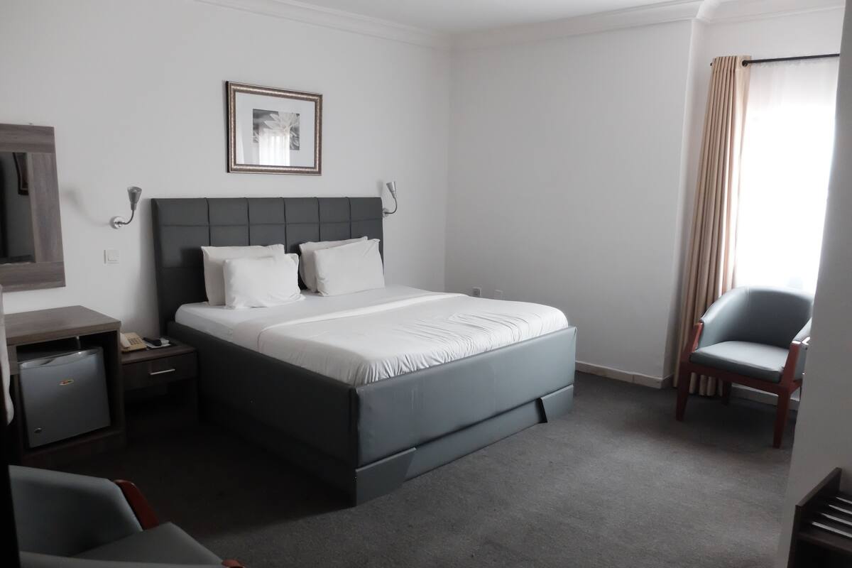 Quarto de hotel com cama, poltrona, escrivaninha e televisão.