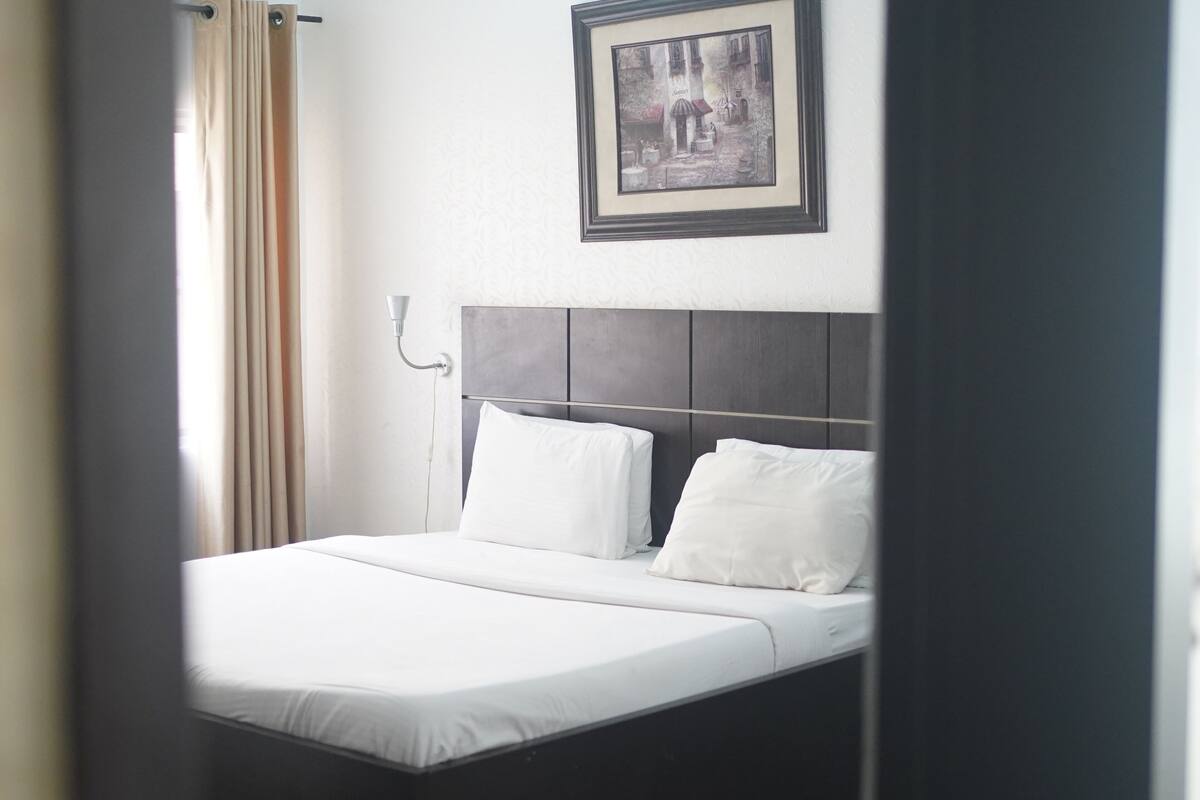 Cama bem arrumada com lençóis brancos e cabeceira escura, um quadro na parede e uma cortina parcialmente aberta.