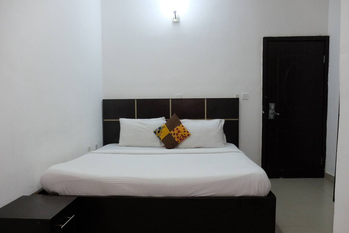 Uma cama com roupa de cama branca e cabeceira preta, um único travesseiro decorativo, uma mesa de cabeceira preta e uma porta fechada.