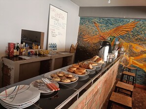 Buffet de café da manhã com diversos alimentos, um espaço para café e um mural na parede.