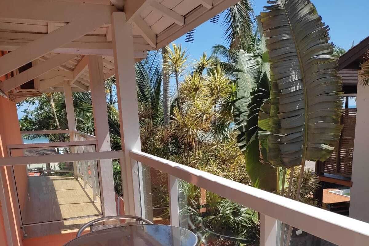 Varanda coberta com mesa e cadeiras de vidro, com vista para um jardim com palmeiras e outras plantas.