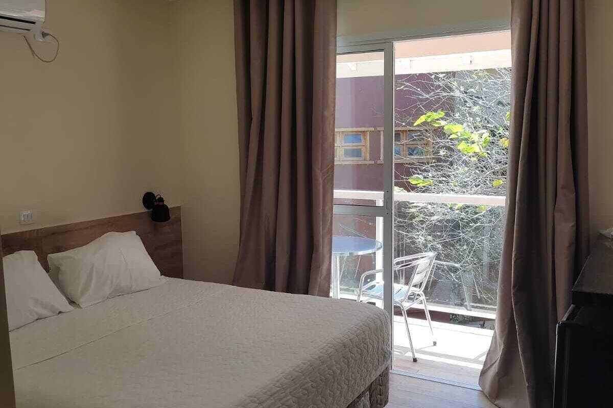 Quarto de hotel com cama, escrivaninha e varanda com cortinas.