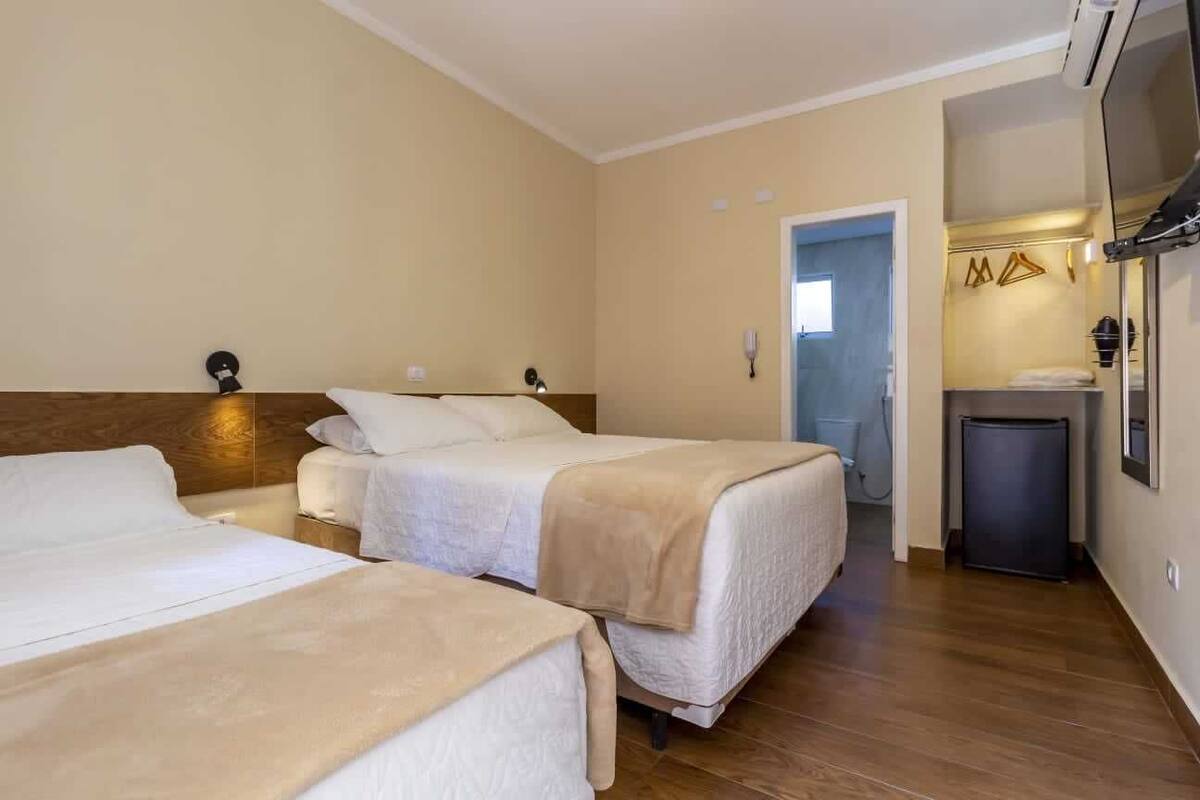 Quarto de hotel com duas camas, cabeceira de madeira, pequena cozinha e televisão.