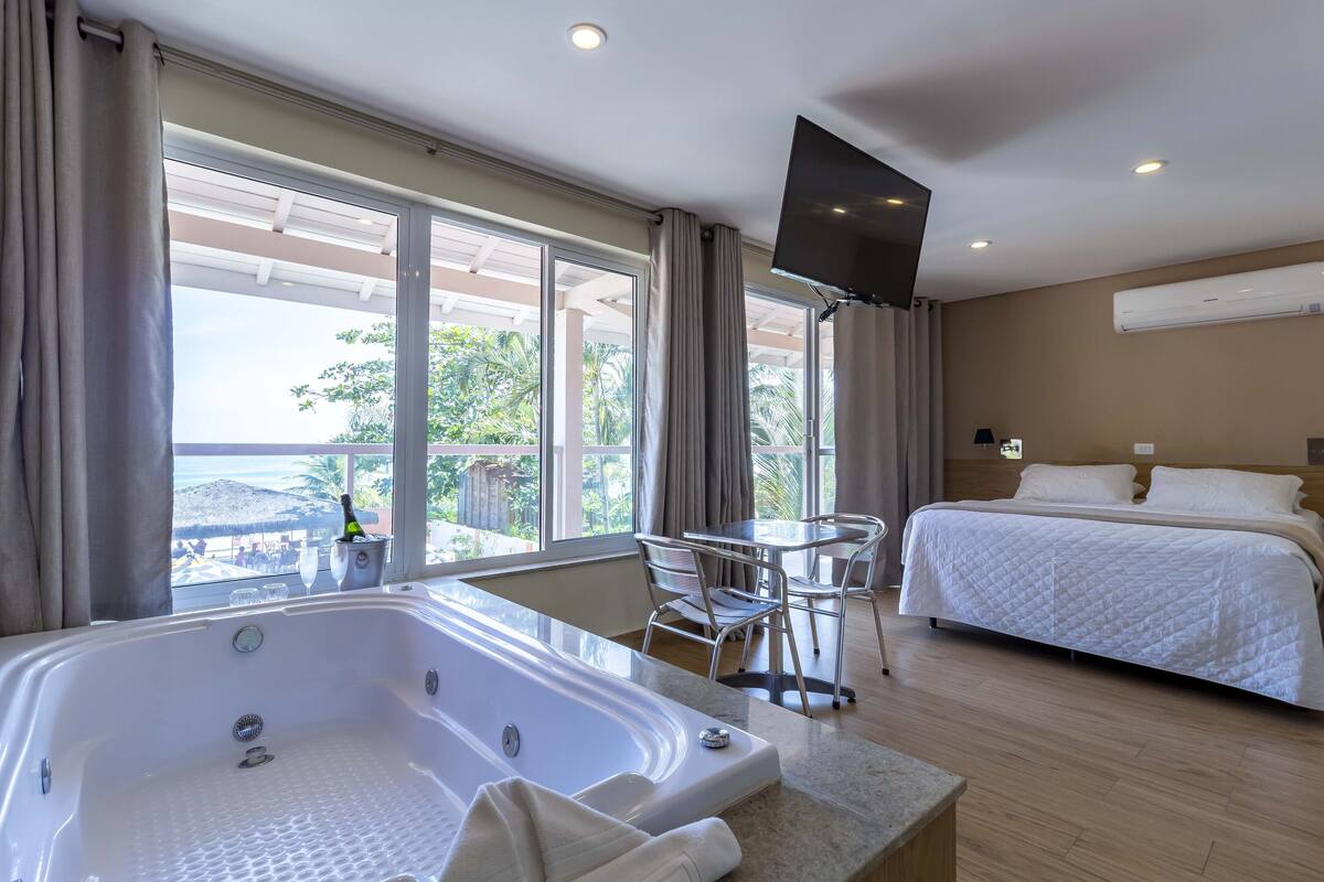 Quarto de hotel com uma janela grande, banheira, cama, televisão e mesa com cadeiras.