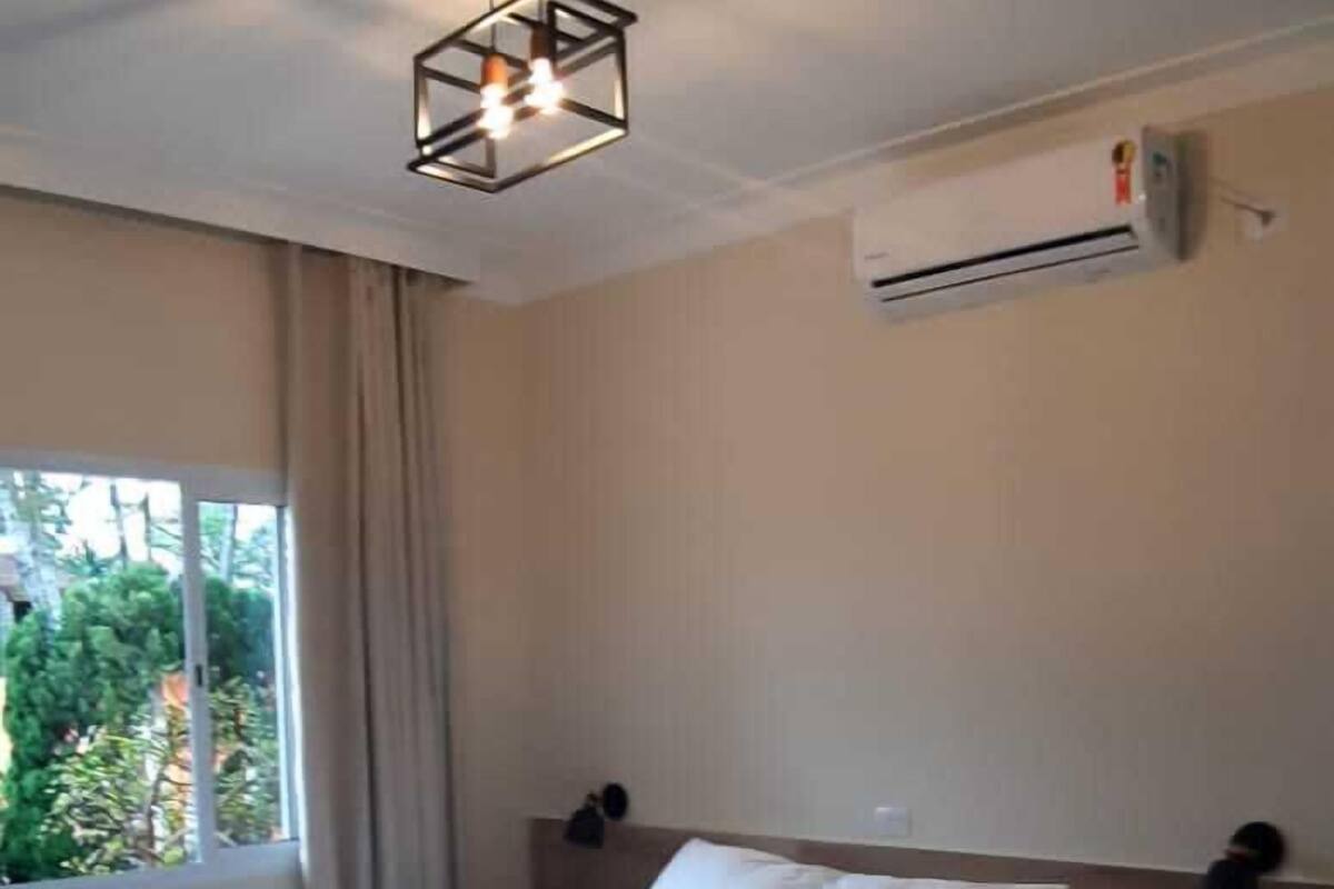 Quarto de hotel com uma cama grande, luminária no teto, aparelho de ar condicionado e uma janela com cortinas.