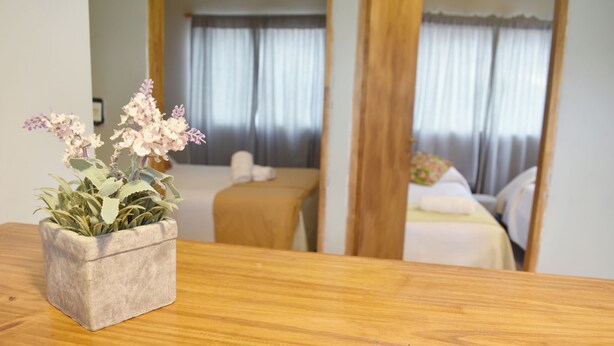 Uma mesa de madeira com uma planta em vaso quadrado. Ao fundo, uma cama com colcha branca e uma janela com cortinas.