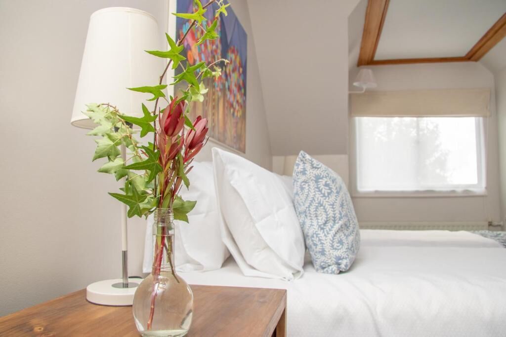 Quarto com cama, mesa de cabeceira, abajur, um vaso com flores e janela com cortinas.