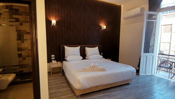 Quarto de hotel com uma cama grande, duas luminárias laterais, cabeceira de madeira e uma varanda com cadeira.