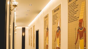 Um corredor com arte nas paredes inspirada no Egito, incluindo figuras de Amum-Ra e Hórus, e um interior moderno e bem iluminado.
