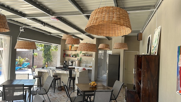 Amplo espaço interno com pé direito alto, vigas aparentes e luminárias de vime pendentes. Há uma área de cozinha com pia, bancada e diversos utensílios. É visível uma área de refeições com mesas e cadeiras, além de um pequeno espaço para sentar ao ar livre com cadeiras e uma mesa.