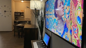 Uma sala de estar moderna com uma grande TV de tela plana na parede, um laptop sobre uma escrivaninha, uma área de refeições com um relógio e uma cozinha ao fundo.