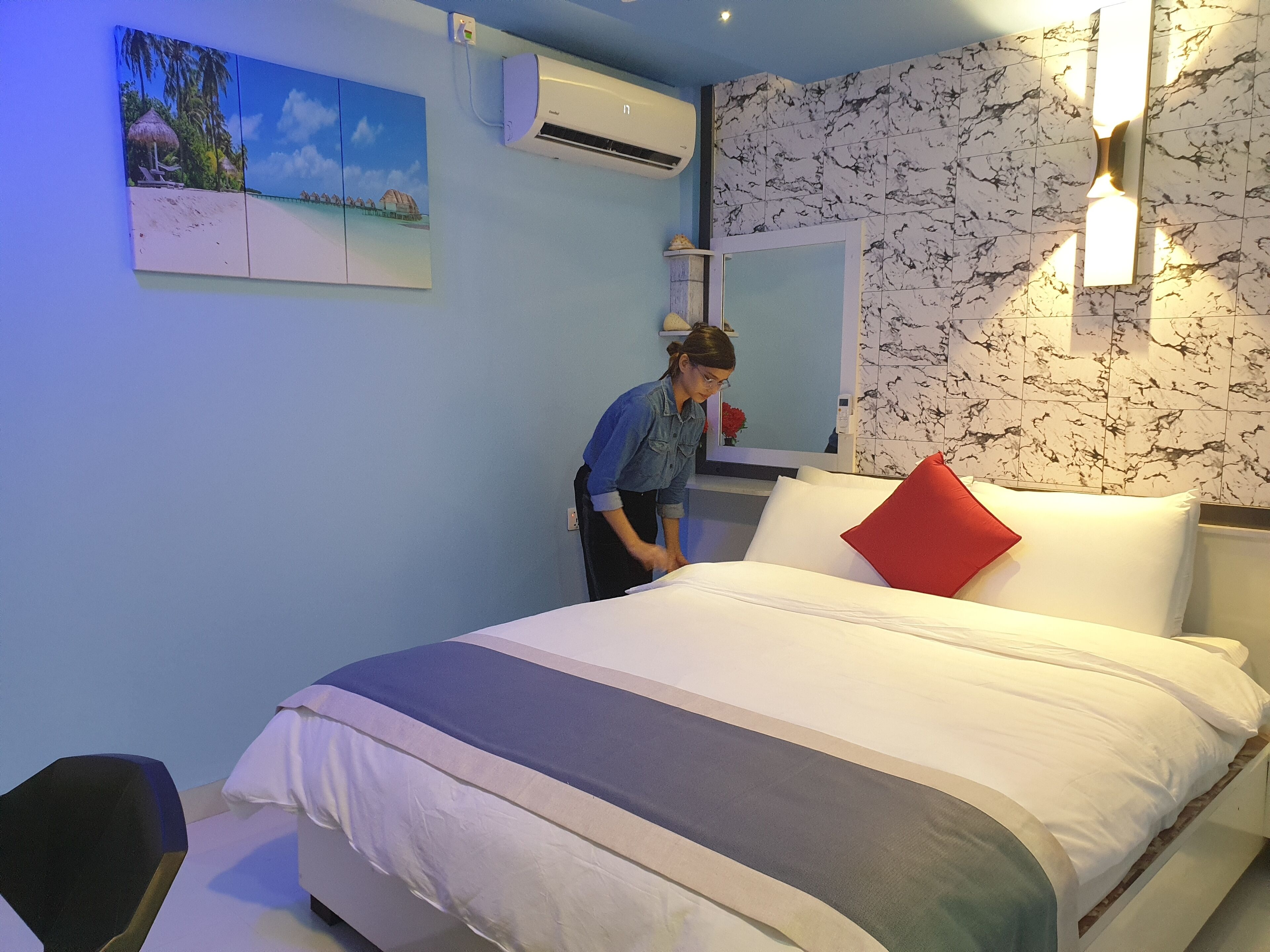 Quarto de hotel com uma cama, uma escrivaninha e uma cadeira. Há uma pintura na parede e um aparelho de ar condicionado.