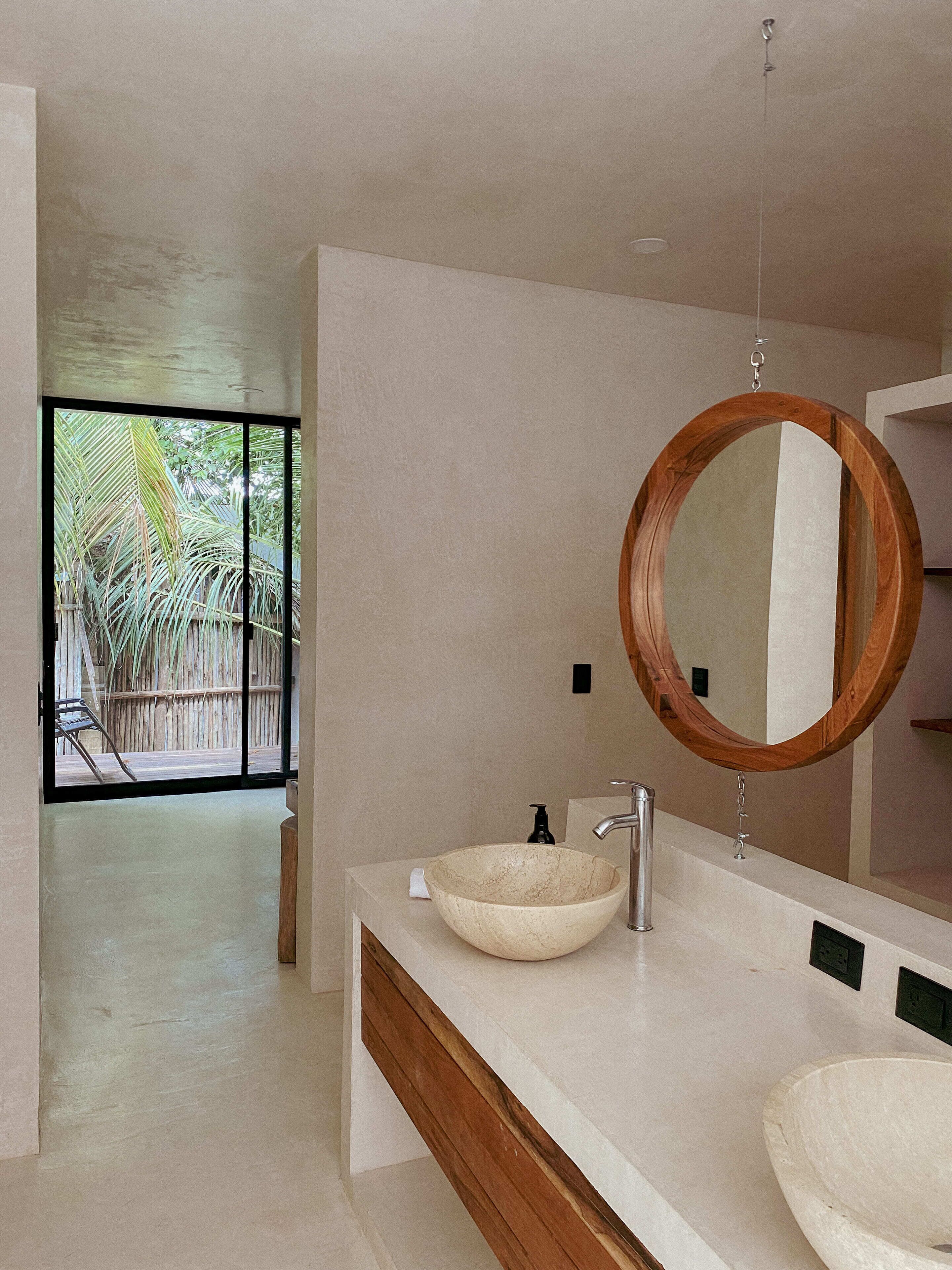 Casa de banho moderna com um espelho grande com estrutura de madeira, duas lavatórios suspensos e vista para uma cerca de bambu através de uma porta de vidro.