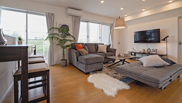 Uma sala de estar moderna com sofá, poltrona e mesa de centro, uma TV na parede e uma varanda com planta.