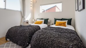 Quarto com duas camas, cada uma com colcha e travesseiros franzidos, uma janela com vista para um edifício e uma mesa de cabeceira com abajur.