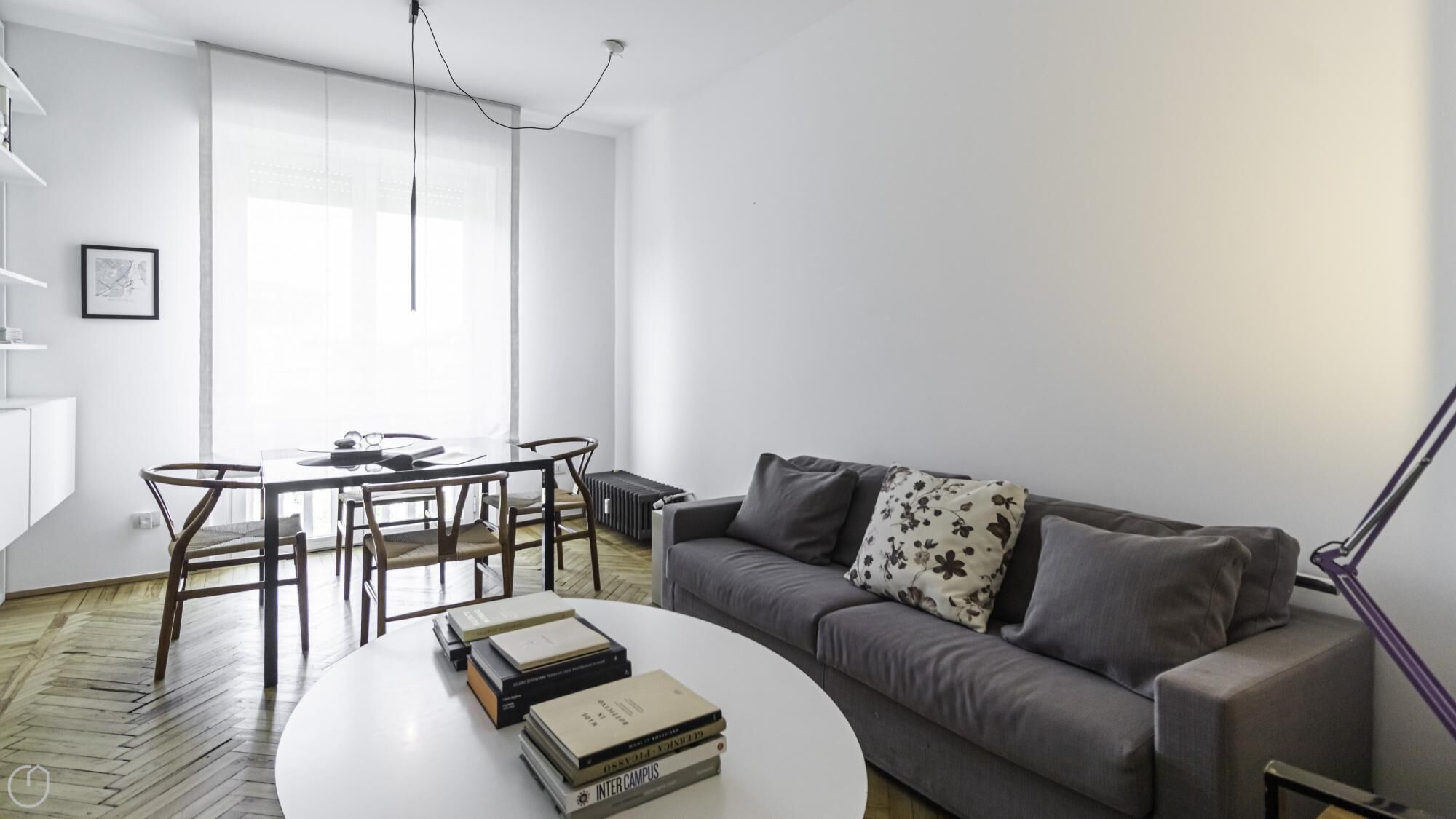 Uma sala de estar moderna com um sofá cinzento, uma mesa de centro redonda com livros, uma mesa de jantar com cadeiras e uma grande janela com cortinas leves.