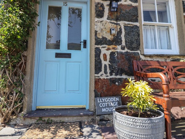 Entrada de uma cabana com porta azul, parede de pedra, um banco e uma placa com a inscrição 'Llygoden Cottage Mouse'.
