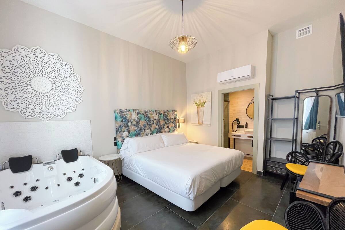Quarto de hotel moderno com uma cama grande, um espaço de estar com uma poltrona amarela, uma banheira branca e um quadro na parede.