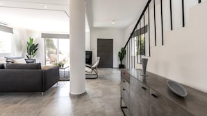 Uma sala de estar moderna com um sofá, uma mesa de centro e uma televisão. Há uma escada com corrimão e uma porta ao fundo.