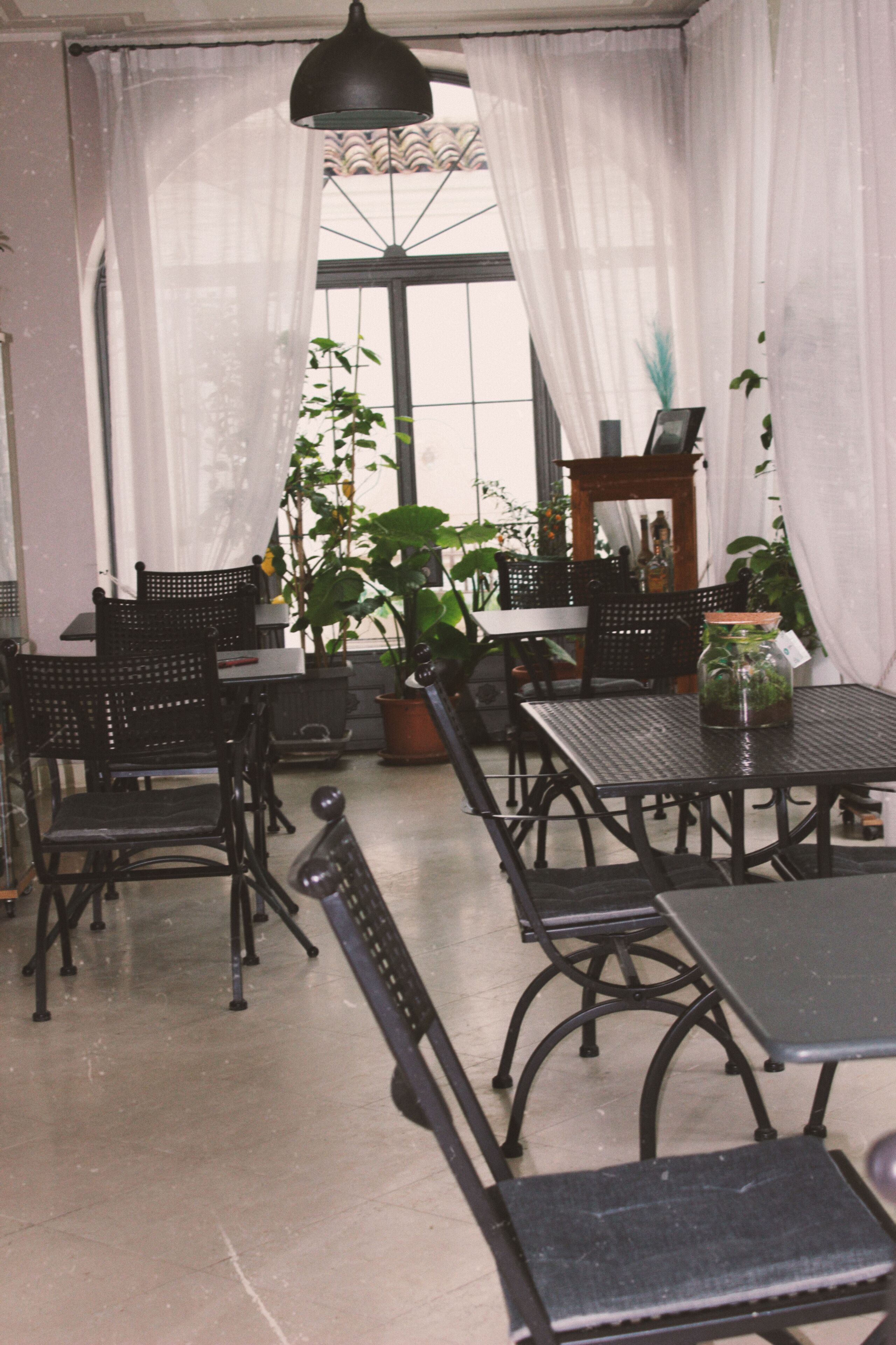 Um quarto com mesas e cadeiras, uma janela grande com cortinas e plantas em vasos.