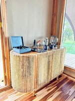 Um aparador de madeira trançada com uma caixa azul contendo canetas e um smartphone, uma jarra de vidro e uma cafeteira em cima.