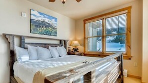 Quarto com estrutura de cama de madeira, uma janela grande com persianas, um quadro na parede e uma mesa de cabeceira com abajur.