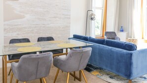 Uma sala de estar moderna com uma mesa de jantar de vidro, cadeiras estofadas cinzas, um sofá azul e um grande mural abstrato na parede.