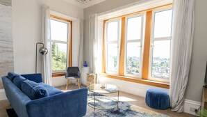Sala de estar com sofá azul, mesa de centro redonda e vista para a paisagem urbana através de grandes janelas.
