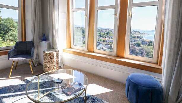 Quarto com janelas amplas, uma cadeira, mesa de vidro e um banquinho azul.