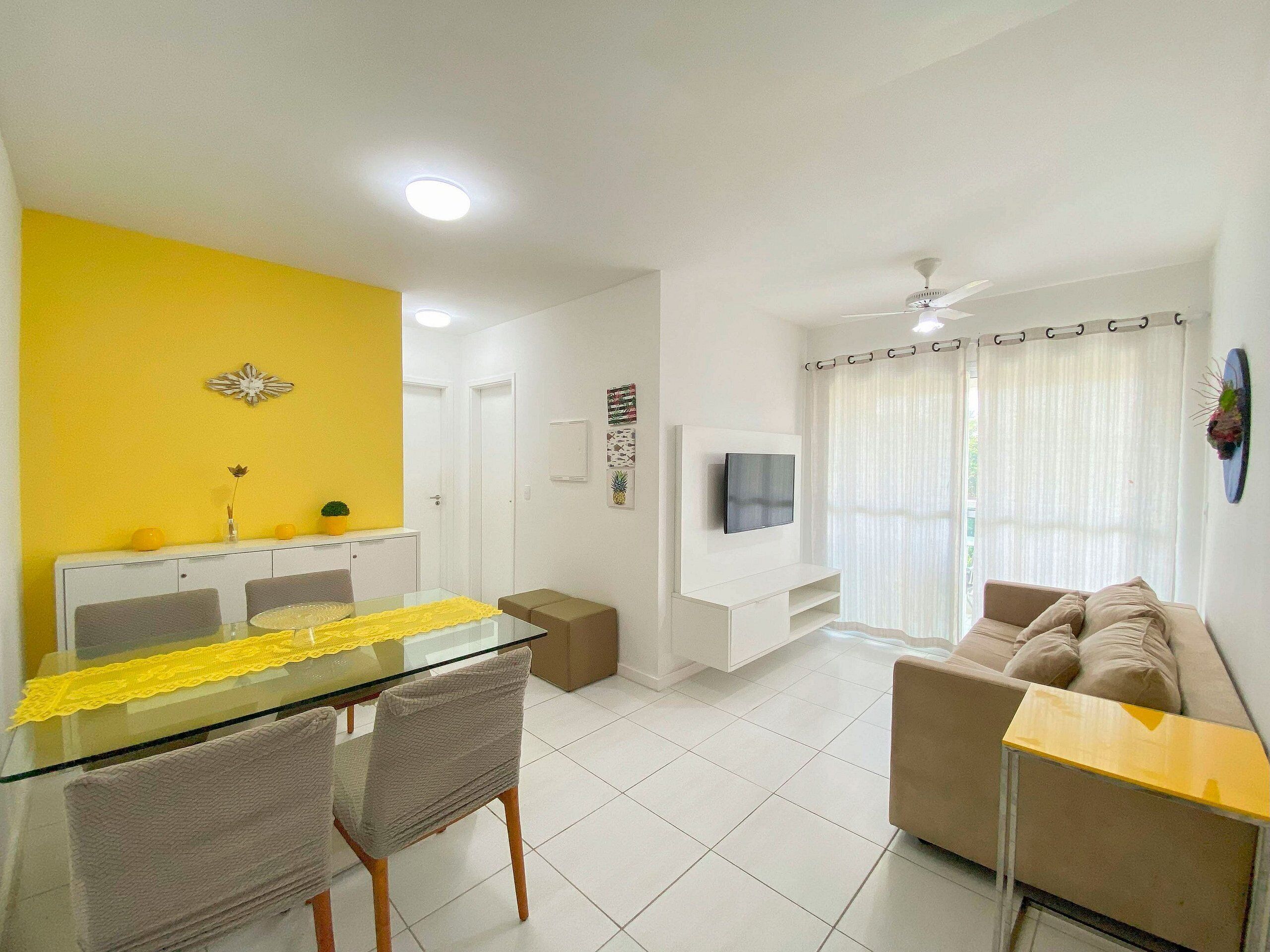 Uma sala de estar com uma mesa de jantar de vidro, um sofá, uma TV fixada na parede e um ventilador de teto.
