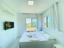 Quarto de hotel com cama, televisão fixada na parede e janela com cortinas.
