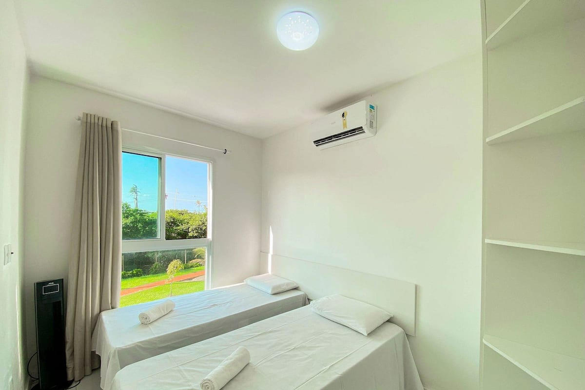 Quarto de hotel com duas camas, ar condicionado, janela com cortinas e vista para o verde lá fora.