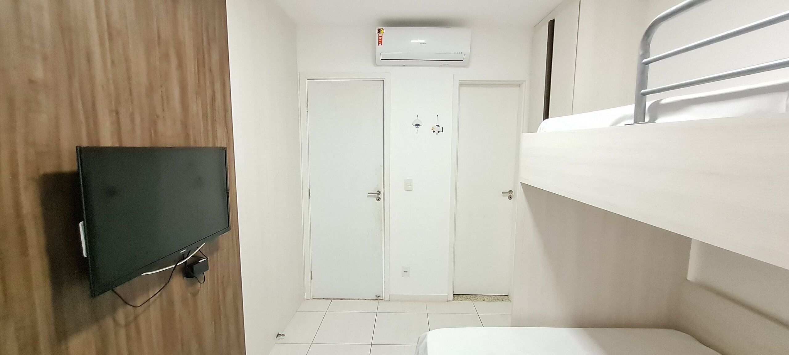 Quarto pequeno e compacto com uma cama de solteiro, uma televisão fixada em um painel de madeira e uma porta com tranca.