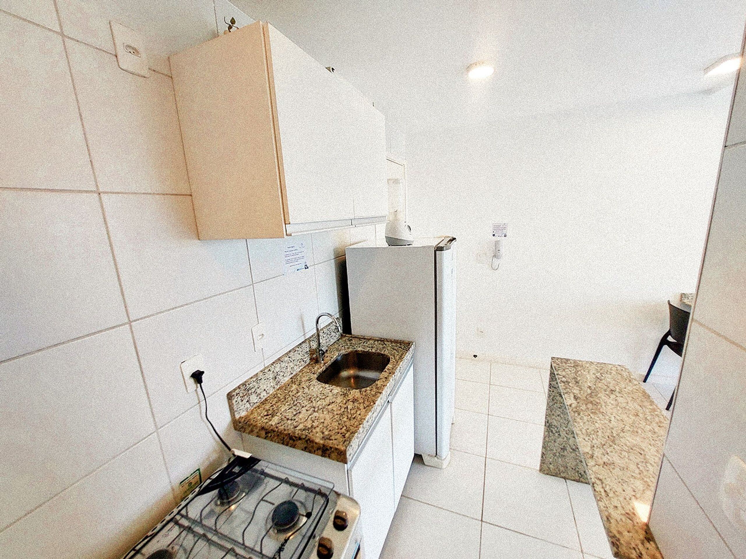 Uma cozinha compacta com paredes revestidas de azulejos brancos, bancada de granito, fogão a gás embutido e geladeira branca.