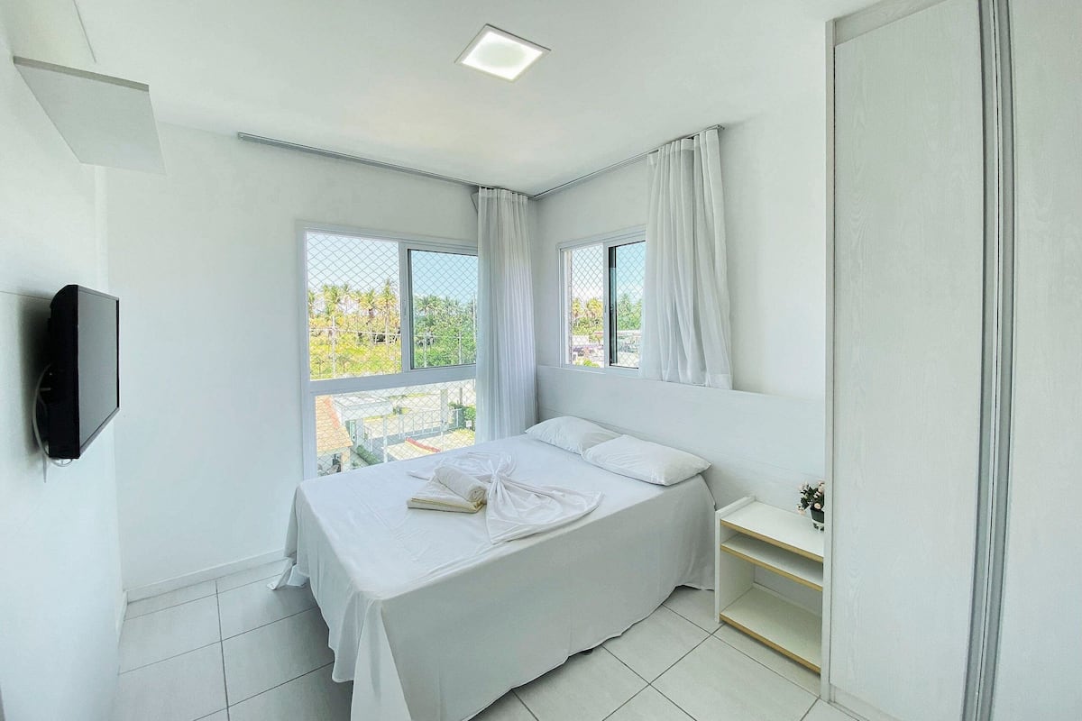 Quarto com cama, mesa de cabeceira, televisão na parede e janela com cortinas.