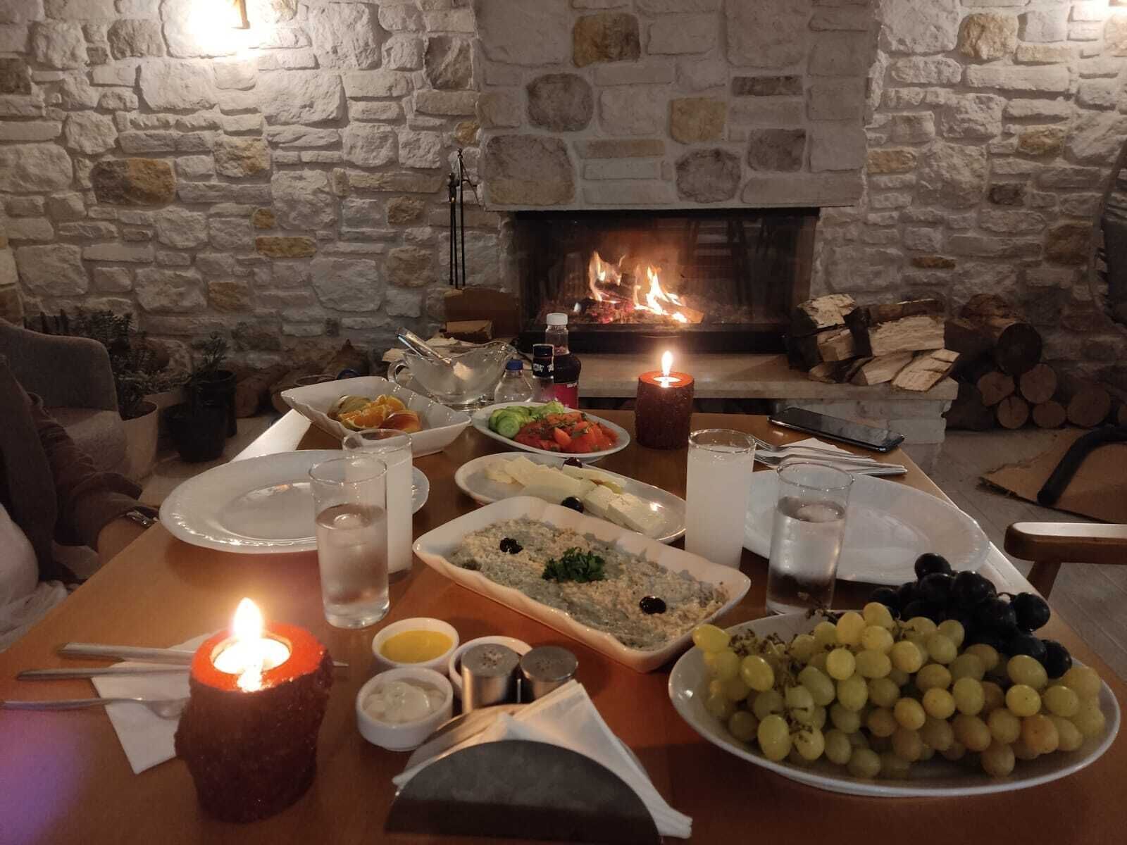 Uma área de refeições acolhedora, com lareira de pedra, uma mesa posta com diversos pratos, copos e uma vela.