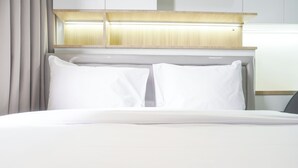 Cama bem arrumada com lençóis e almofadas brancas, cabeceira com iluminação integrada e ambiente em tons neutros.