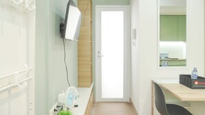 Quarto de hotel com escrivaninha de madeira, uma cadeira, pequeno ventilador, garrafa de água e espelho na parede.