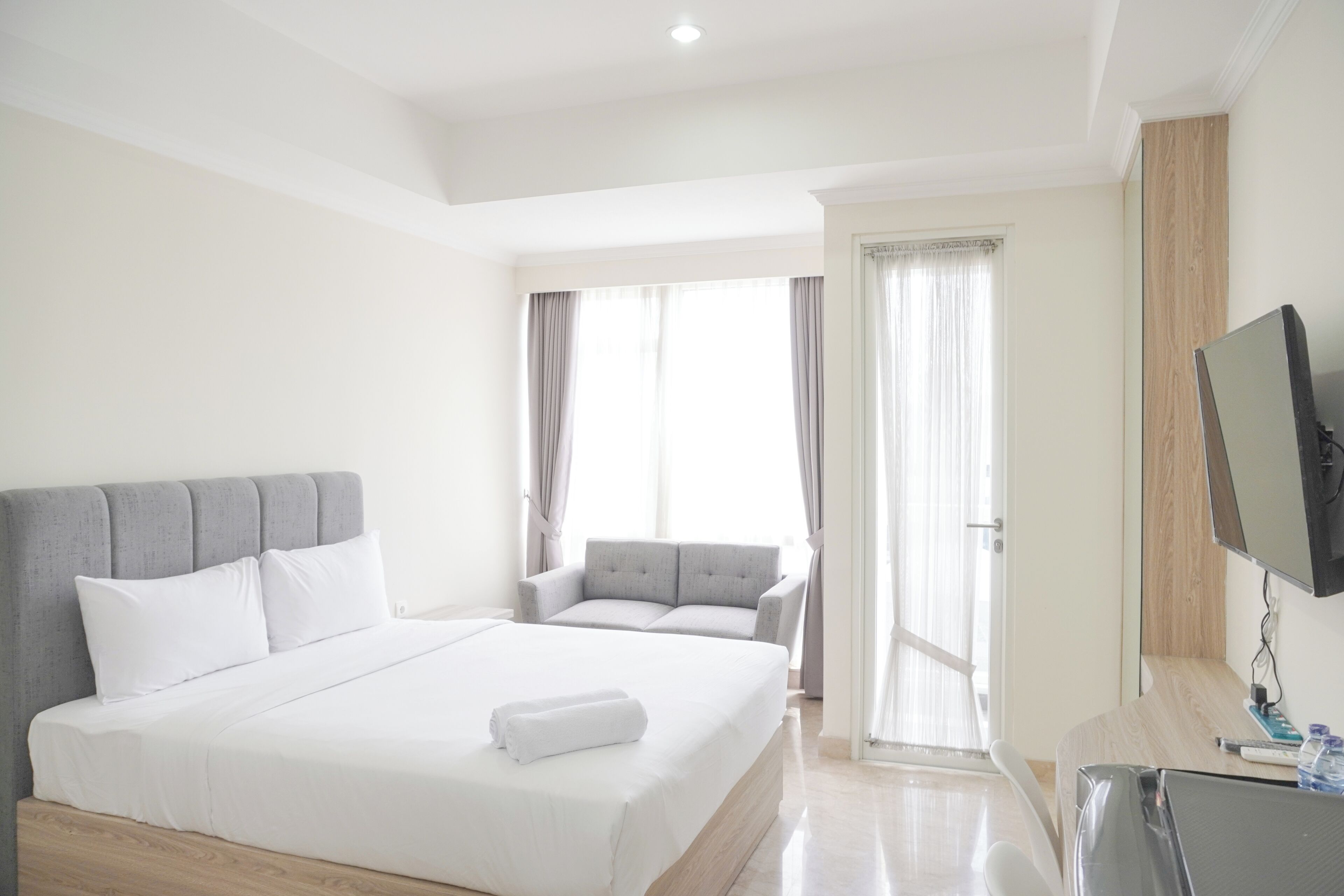 Quarto de hotel com uma cama grande, um sofá cinza estofado, uma televisão e uma janela com cortinas.