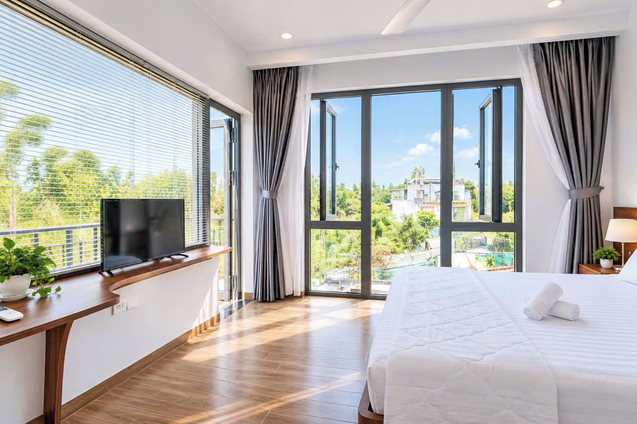 Quarto de hotel com cama, televisão sobre um console de madeira, janela com cortinas e vista para o verde lá fora.