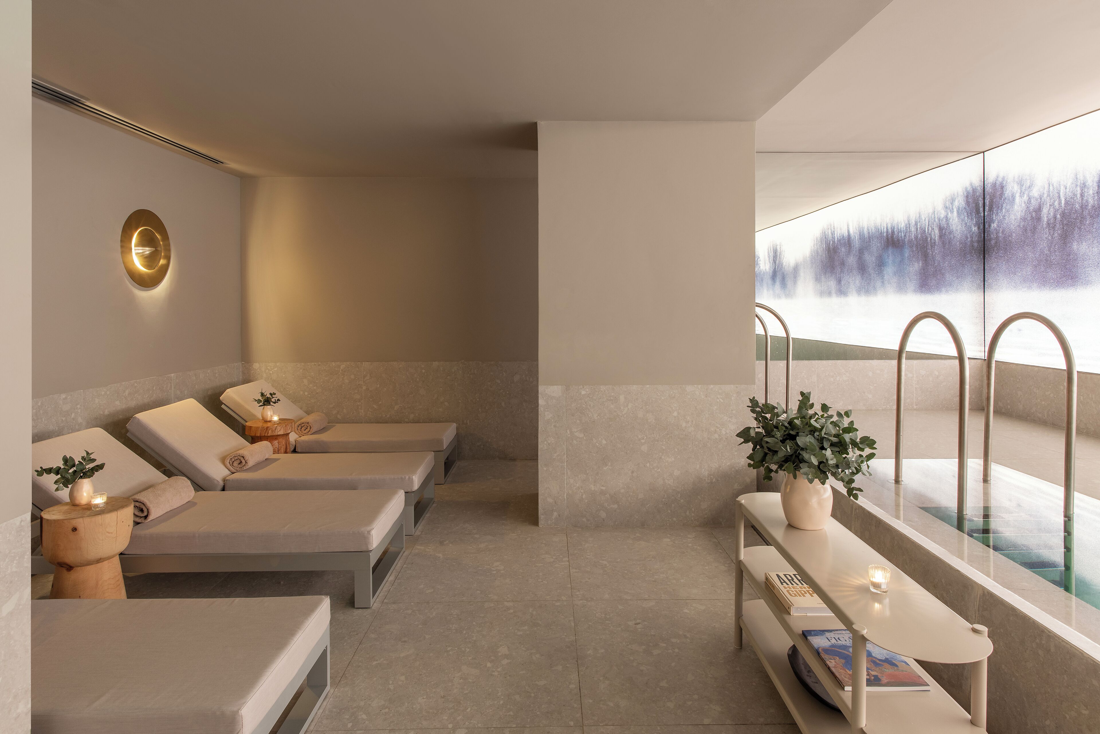 Uma área de spa com três macas de massagem, um banco e uma pequena mesa com uma planta e velas.