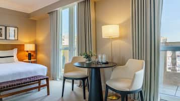 Quarto de hotel com cama, duas poltronas, uma mesinha, luminária e janela com cortinas.